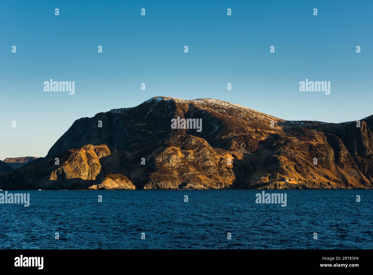 Steep cliff on the coast with small houses and snow on top in Norway on ...