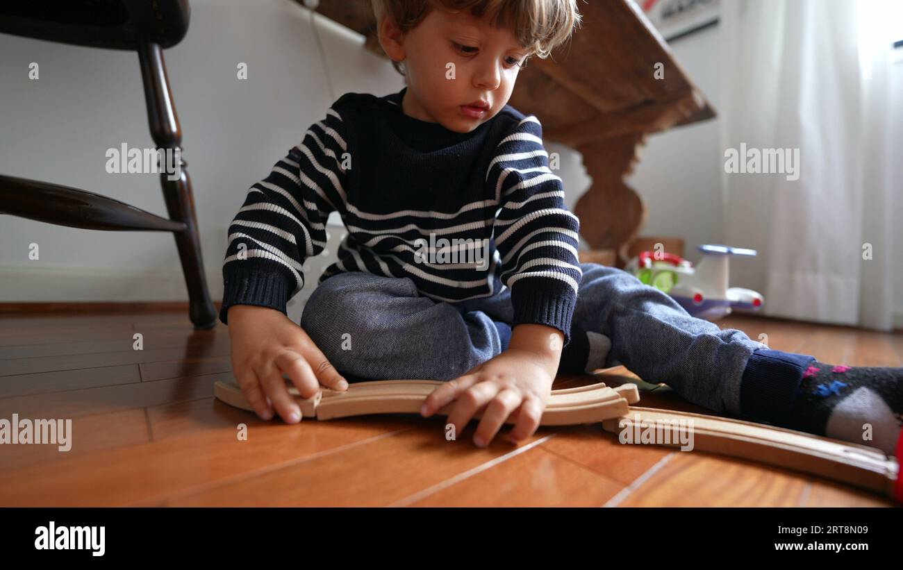 Small boy seated on hardwood floor putting wooden train tracks together ...