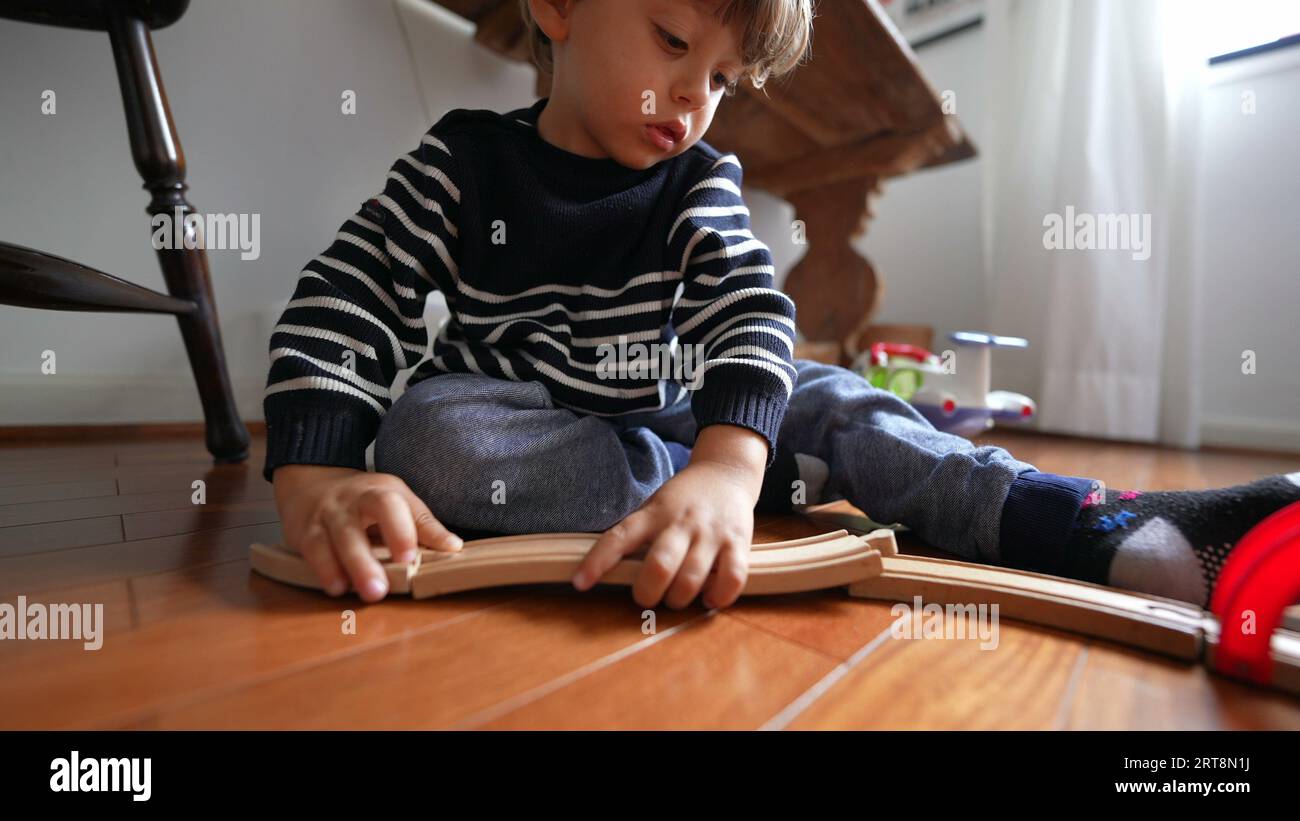 Small boy seated on hardwood floor putting wooden train tracks together ...
