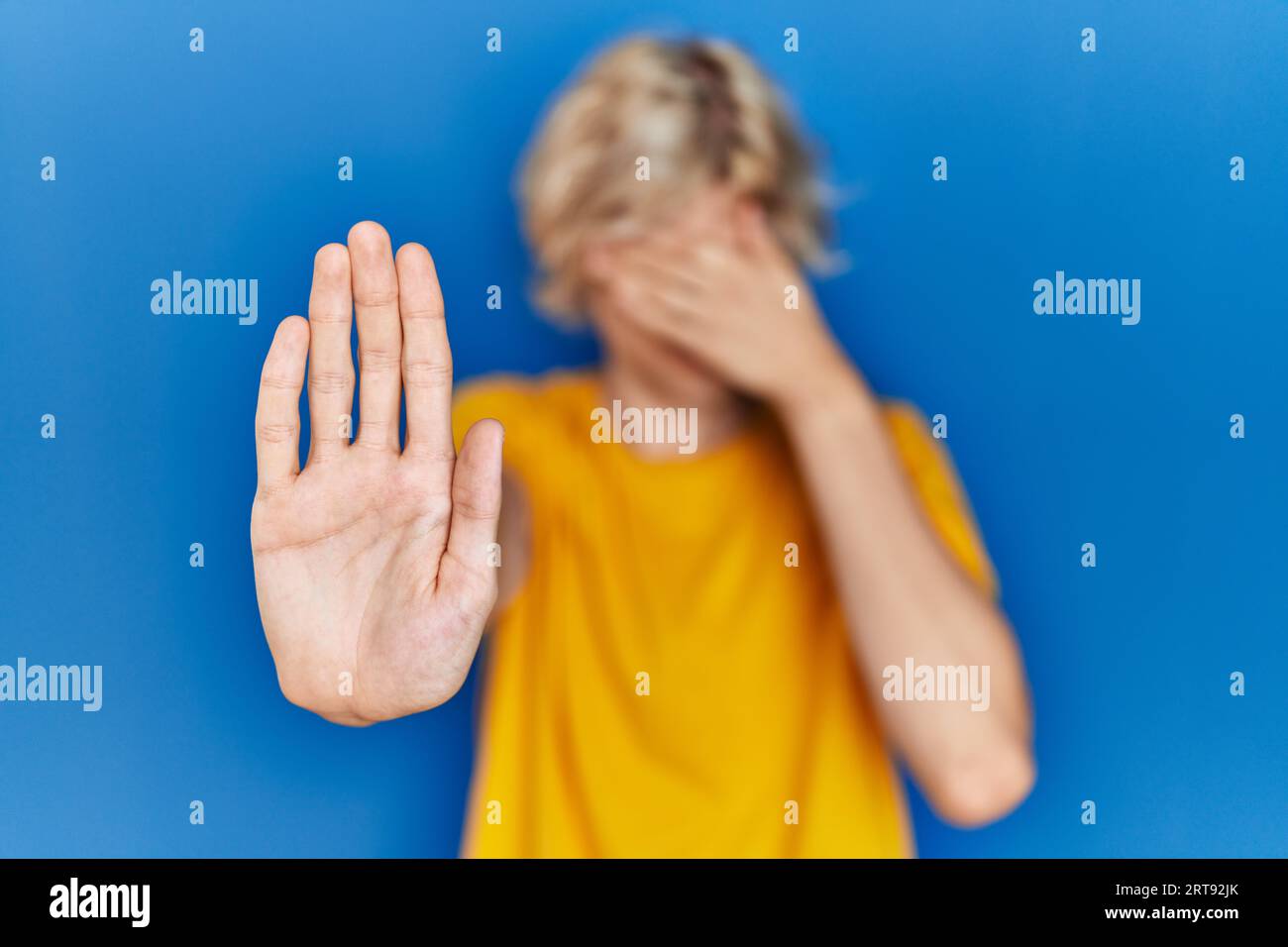 Young modern man standing over blue background covering eyes with hands ...