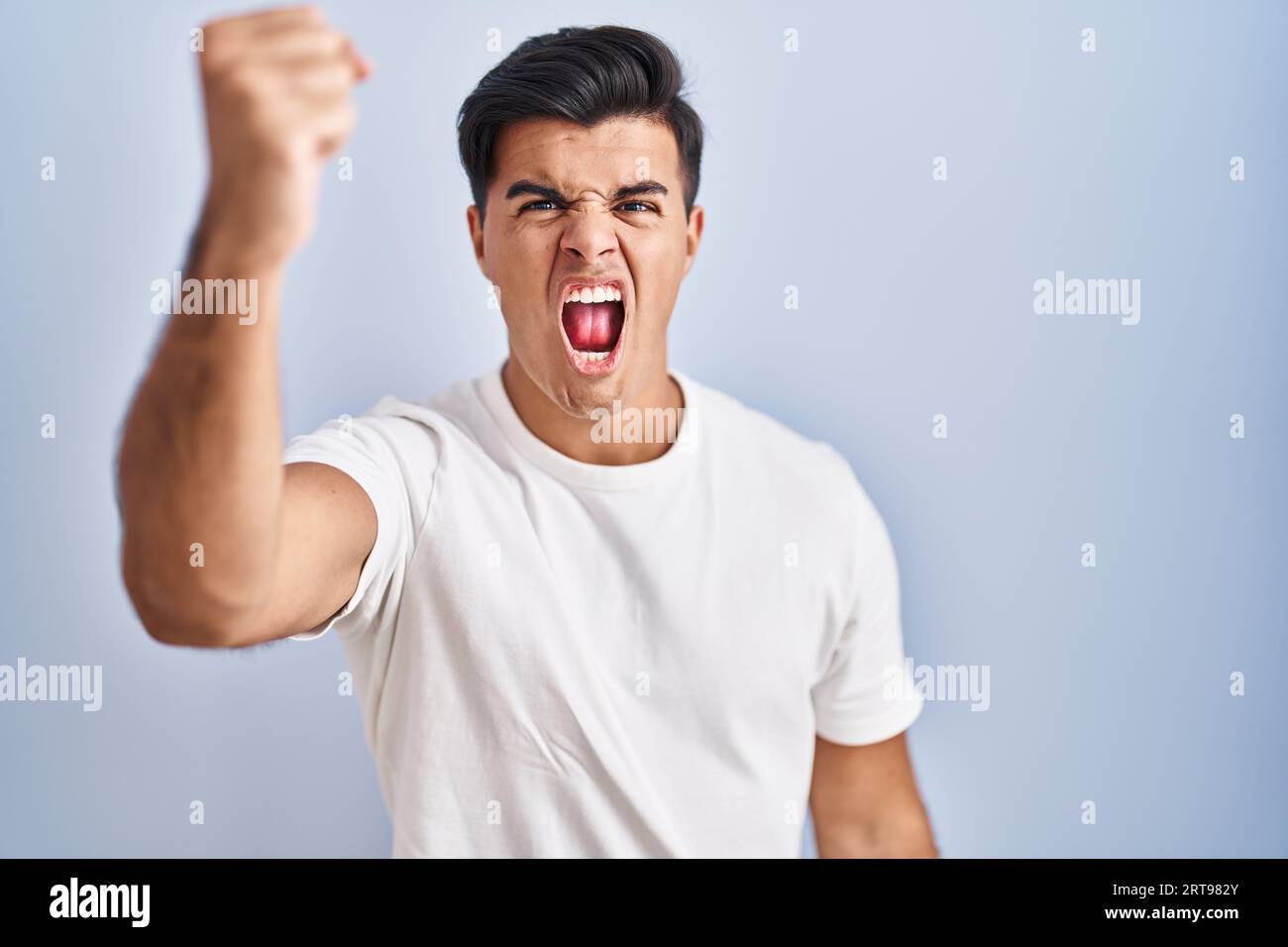 Hispanic man standing over blue background angry and mad raising fist ...