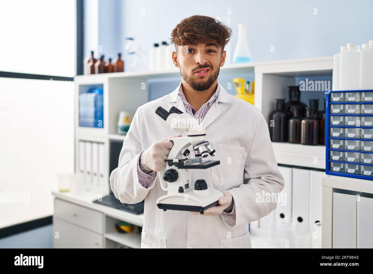 Arab man with beard working at scientist laboratory holding microscope clueless and confused ...