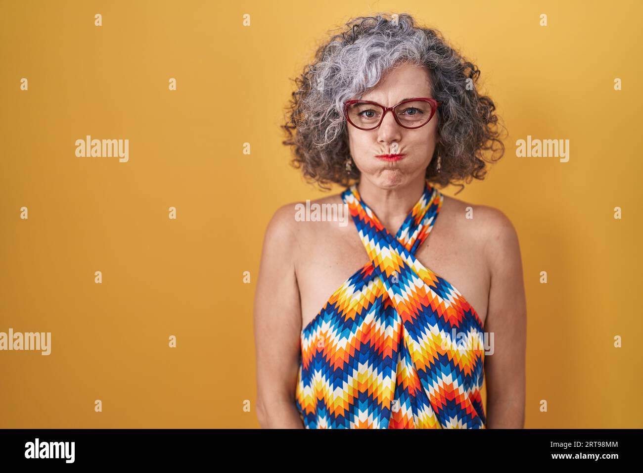 Middle age woman with grey hair standing over yellow background puffing ...