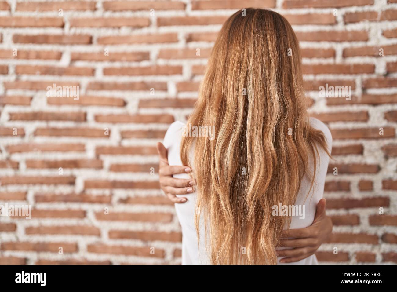 Young caucasian woman standing over bricks wall hugging oneself happy ...