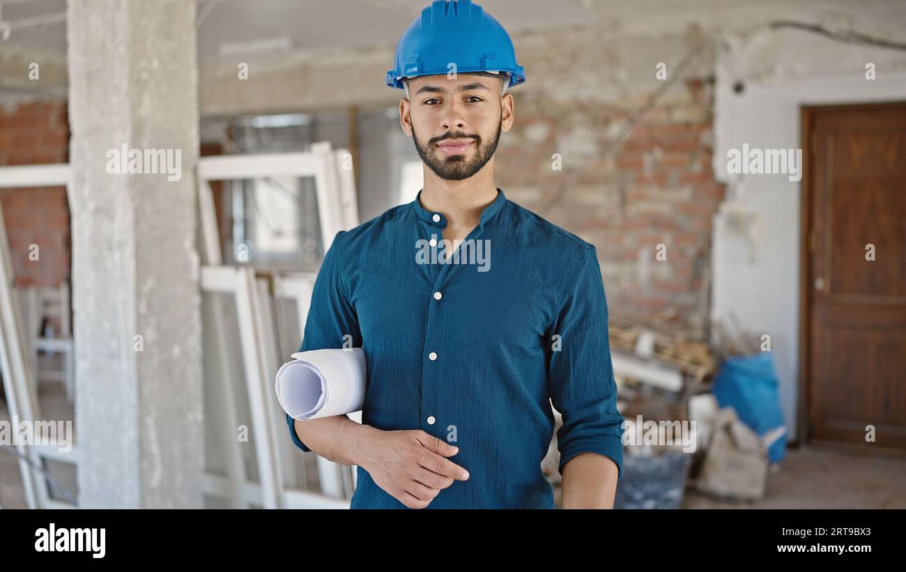Young hispanic man architect wearing hardhat holding blueprints at ...