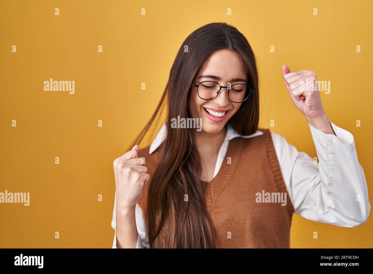 Young brunette woman standing over yellow background wearing glasses ...