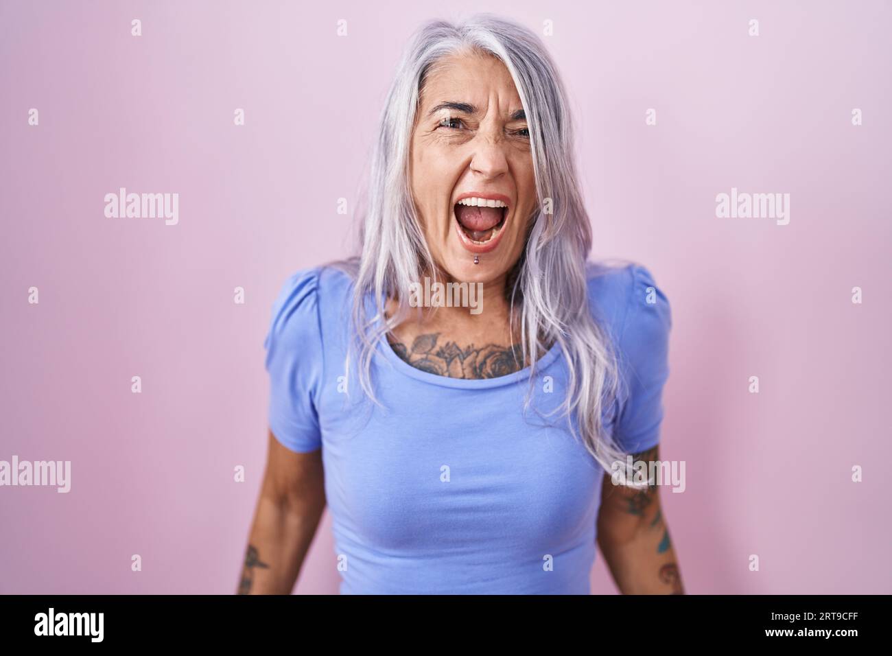 Middle age woman with tattoos standing over pink background angry and ...