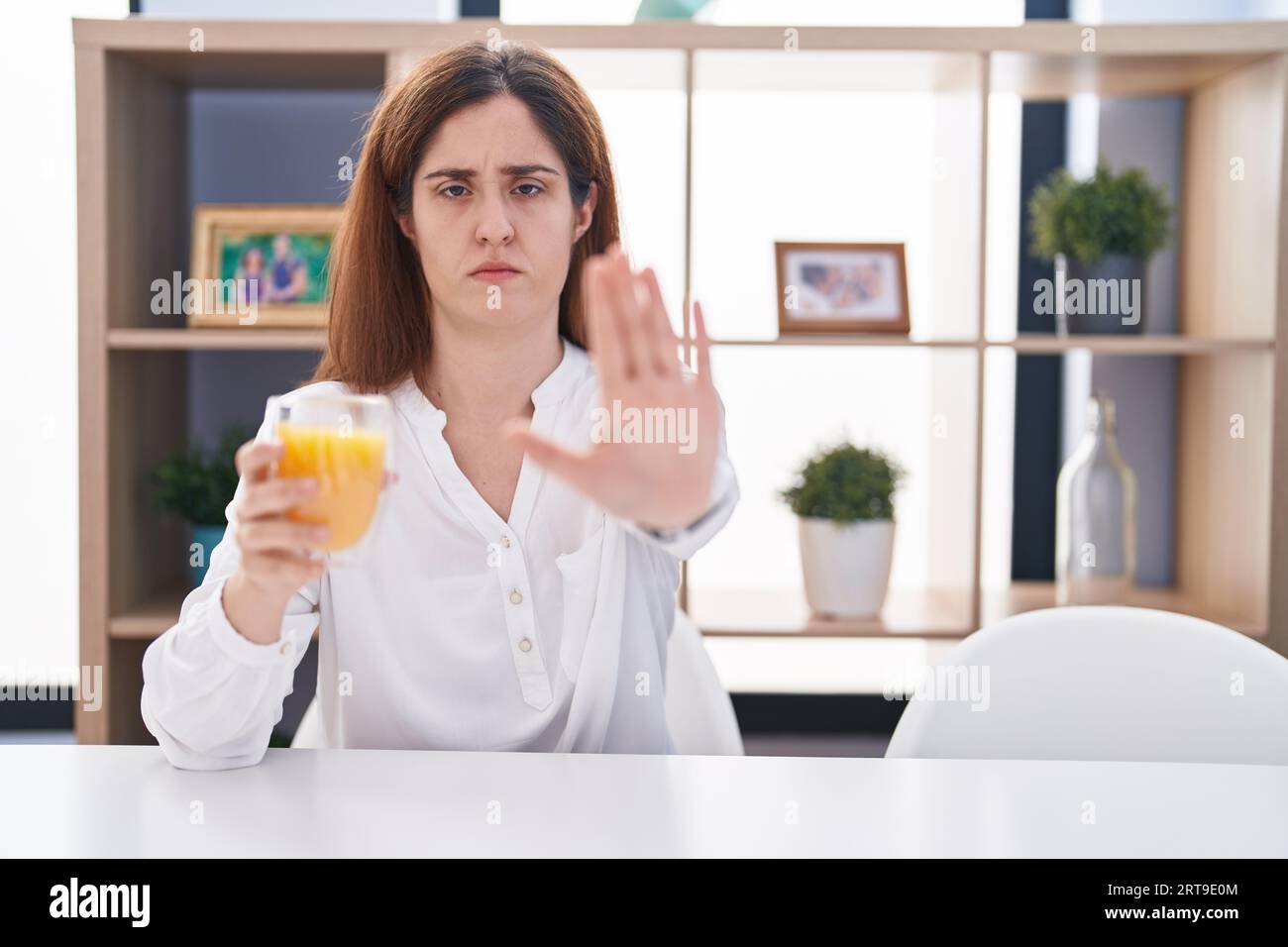 Brunette woman drinking glass of orange juice doing stop sing with palm ...