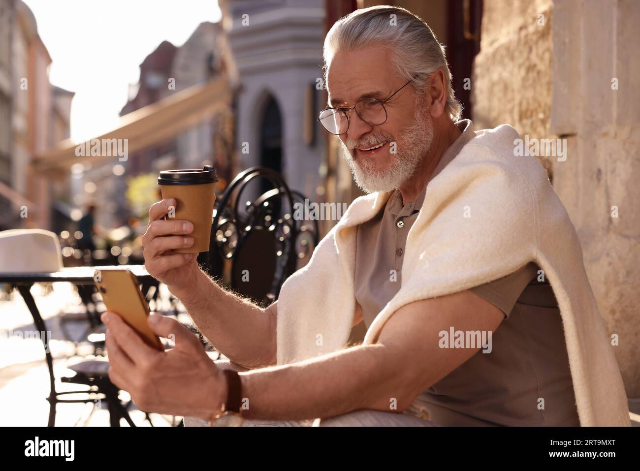 Handsome senior man sitting on doorstep, using smartphone and drinking ...