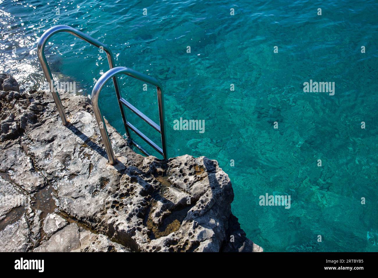 Stainless steel ladder on rock on sea coast. Blue transparent water in ...