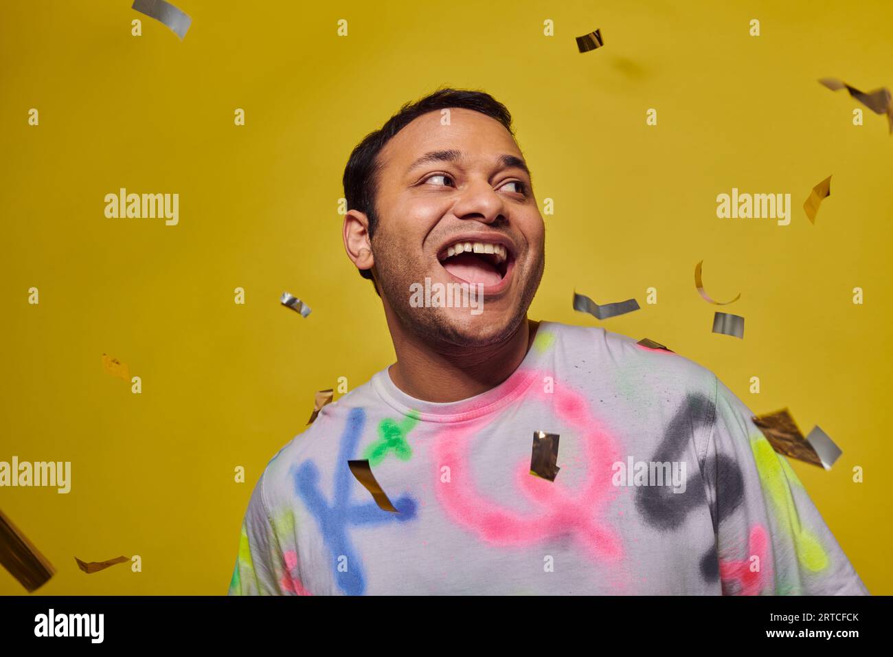 excited indian man smiling near falling confetti on yellow backdrop ...
