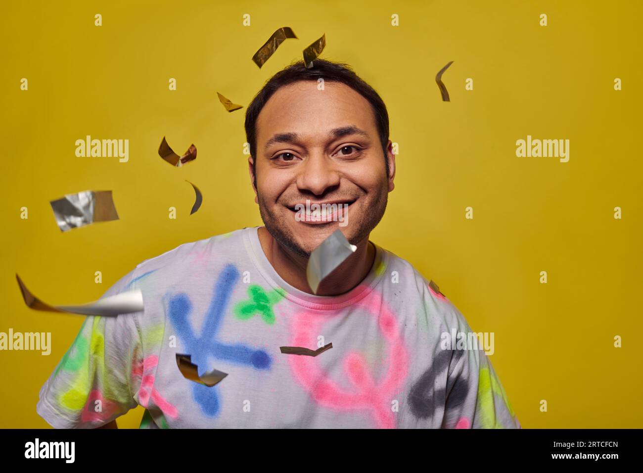 positive indian man smiling near falling confetti on yellow backdrop ...