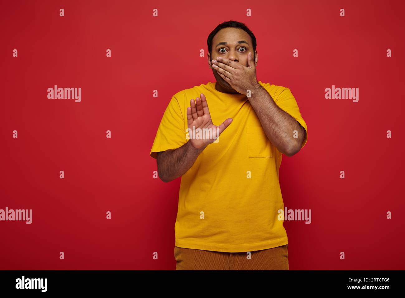 shocked indian man in bright clothes covering mouth and showing stop ...