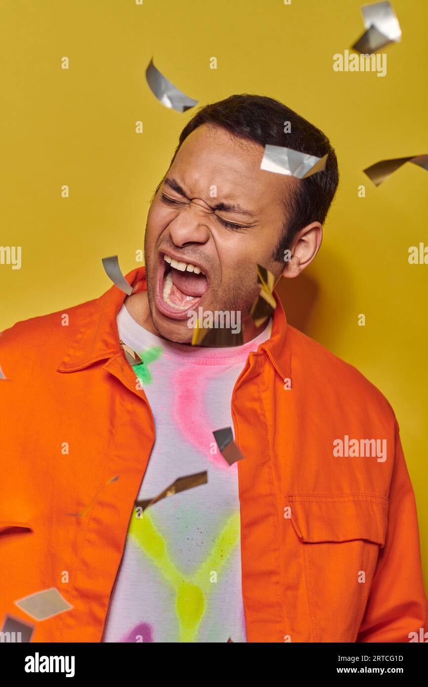 emotional indian man in orange jacket screaming near confetti on yellow ...