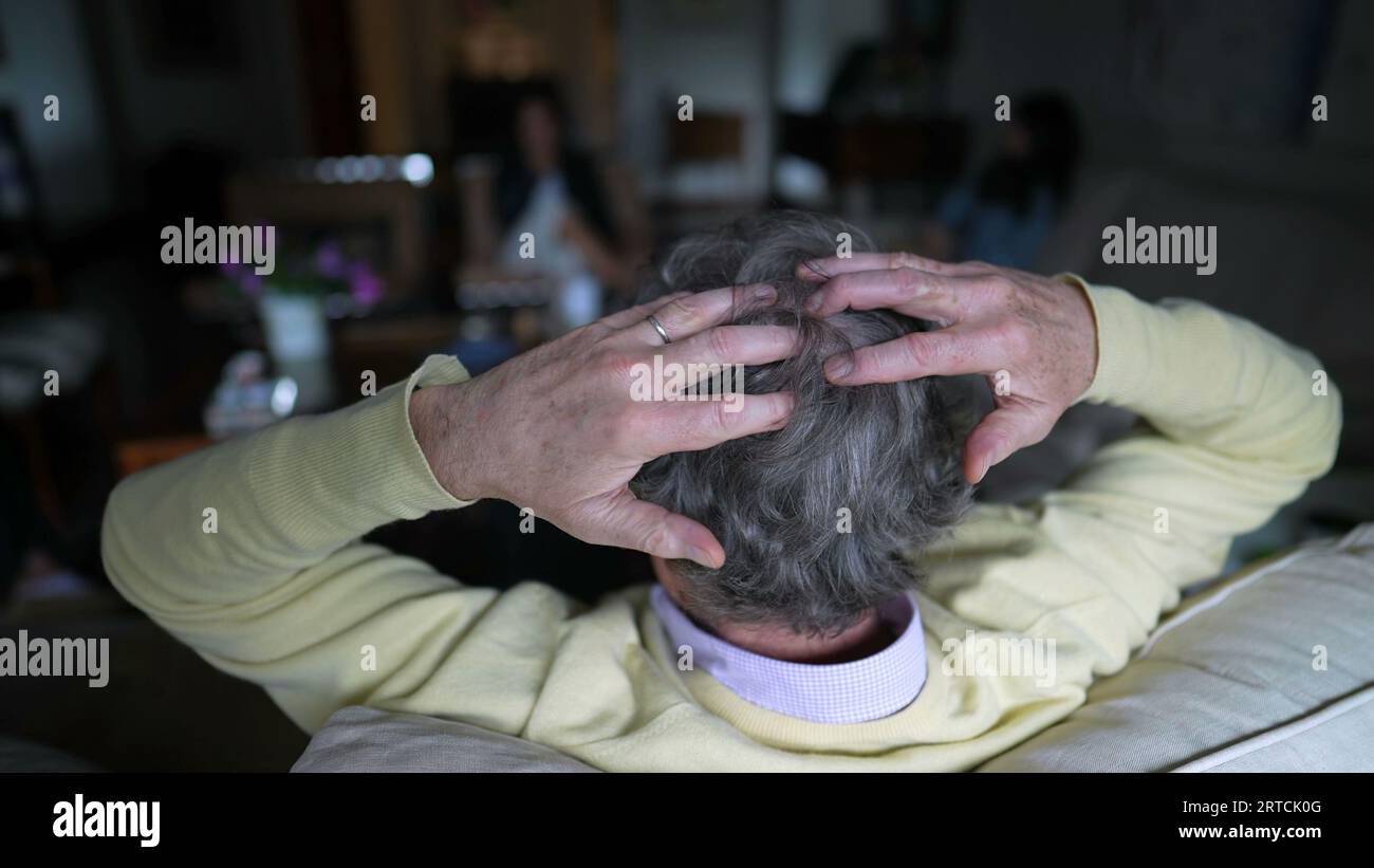 Back of older man scratching head with hands while seated on couch ...
