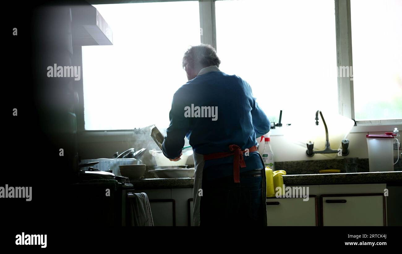 Back of senior chef standing by kitchen sink pouring food into plate ...