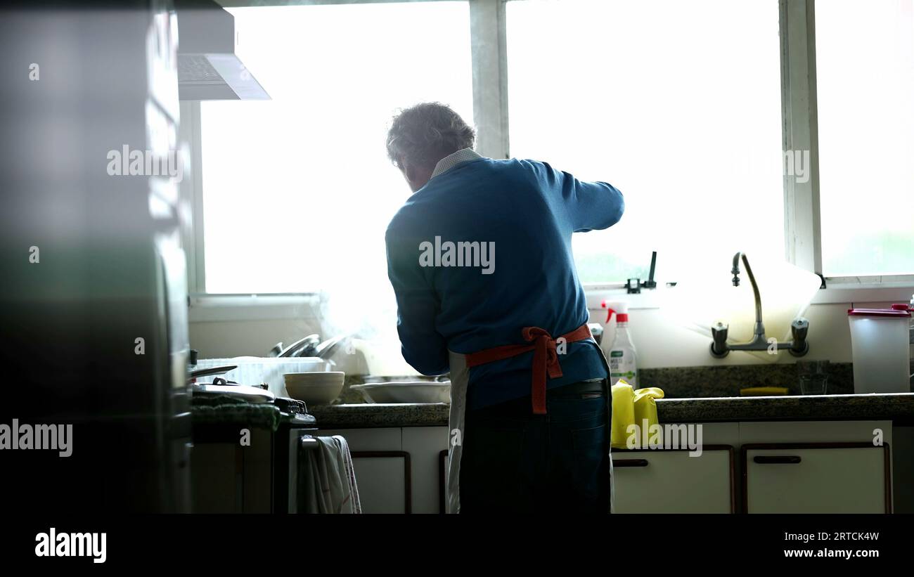 Back of senior chef standing by kitchen sink pouring food into plate ...