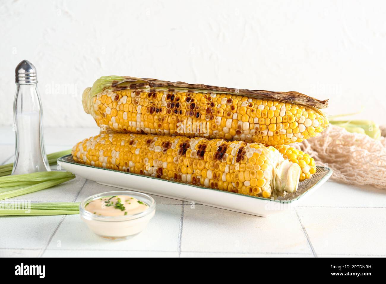 Plate with tasty grilled corn cobs and bowl of sauce on white tile ...