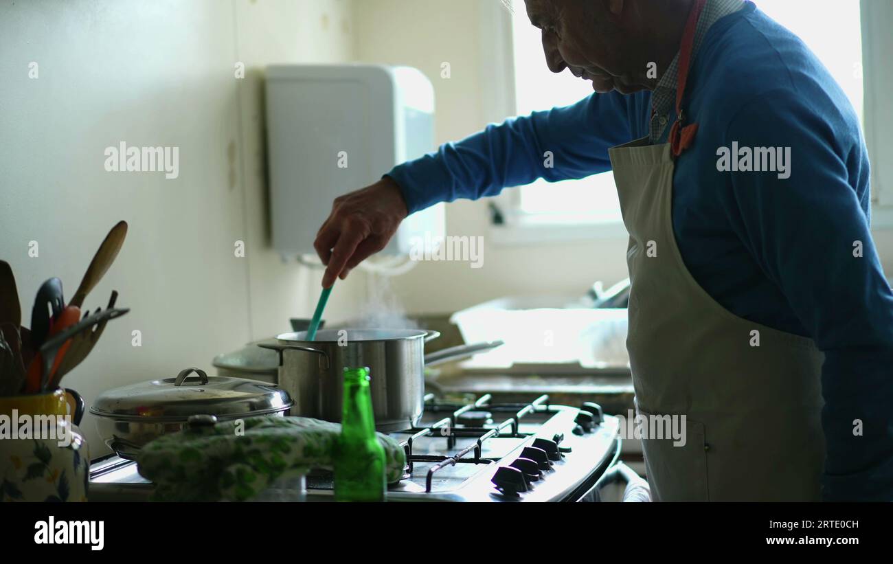 Senior man sipping beer from bottle while cooking food by kitchen stove ...