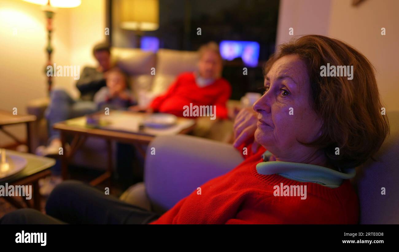 Senior woman watching television seated on couch, family in background ...