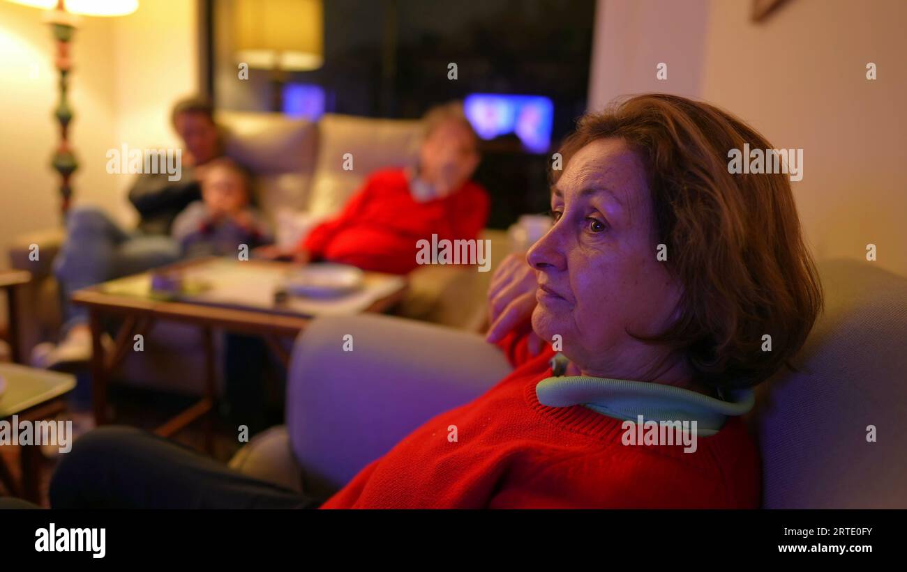 Senior woman watching television seated on couch, family in background ...