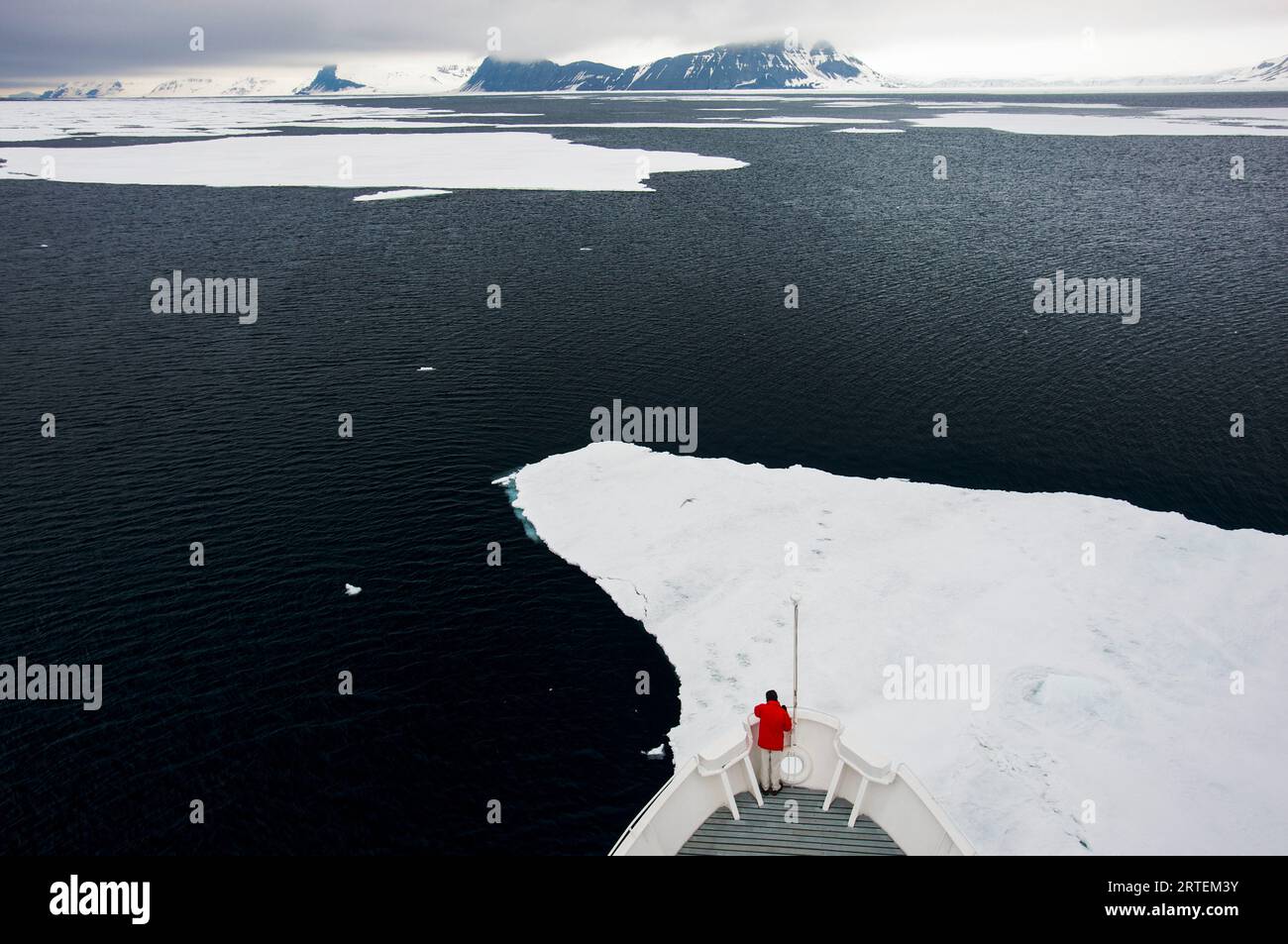 Tourist in a red coat stands looking out from the bow of a cruise ship ...
