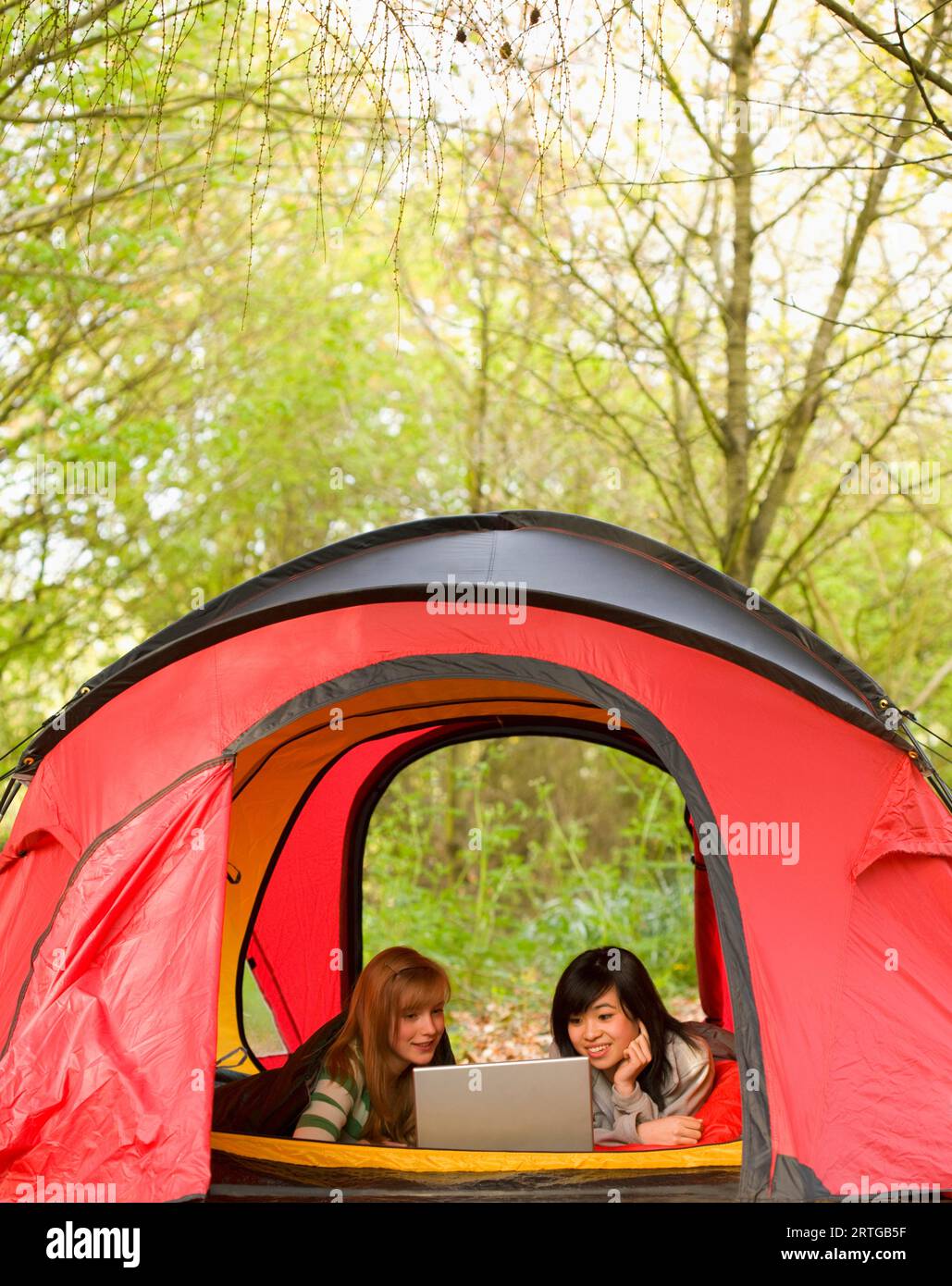 Two smiling teenaged girls lying in a tent looking at laptop computer ...