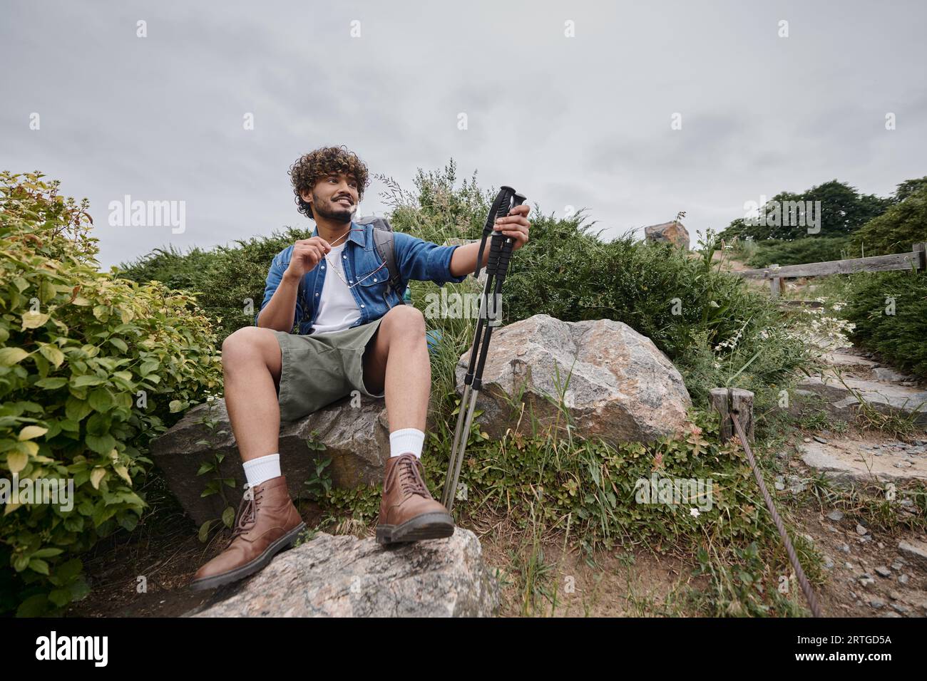 happy indian man sitting with backpack on rock and holding hiking ...