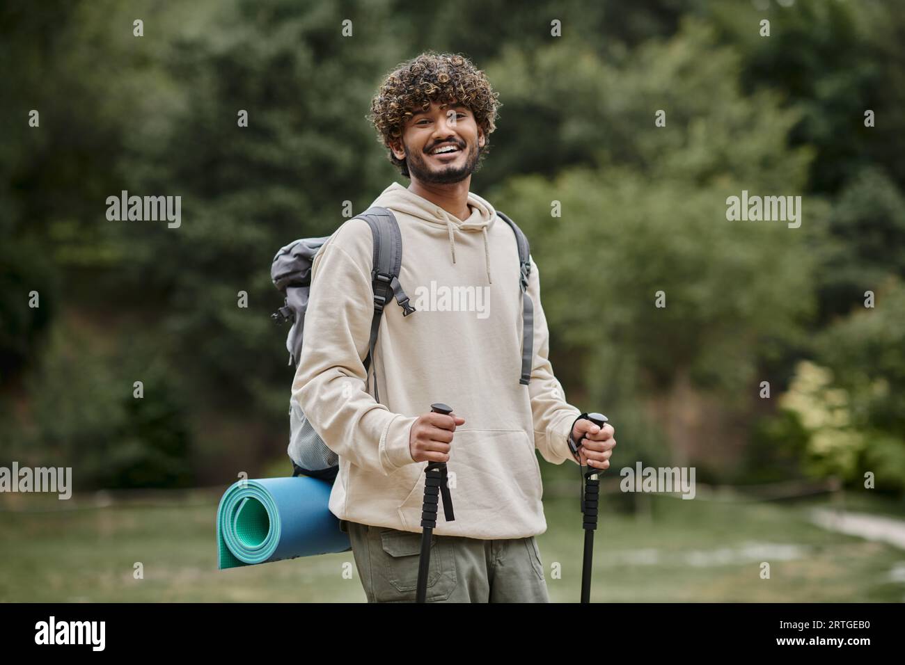 positive indian backpacker holding trekking poles on path in forest ...