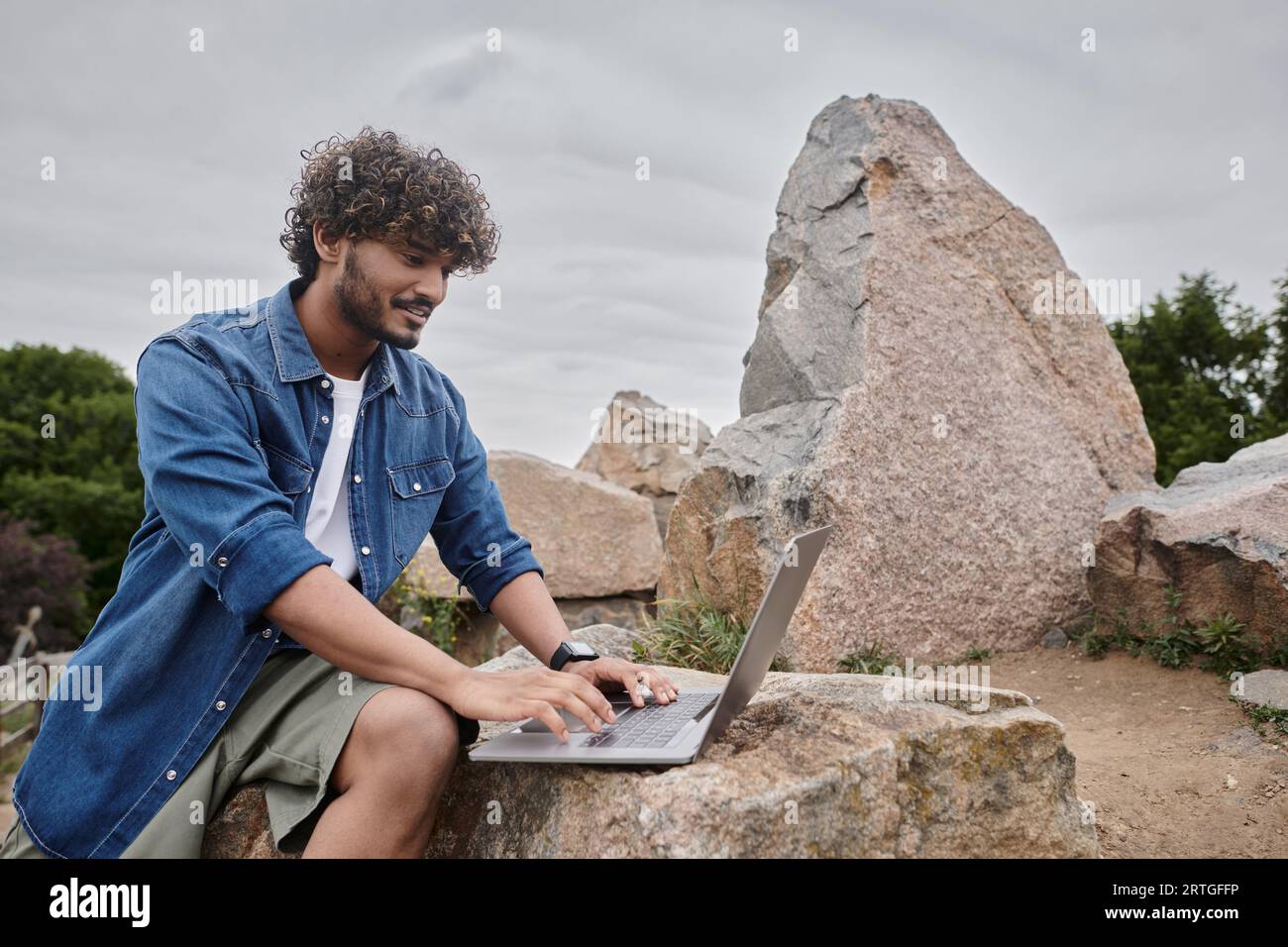 happy indian man typing on laptop and working remotely in natural place ...