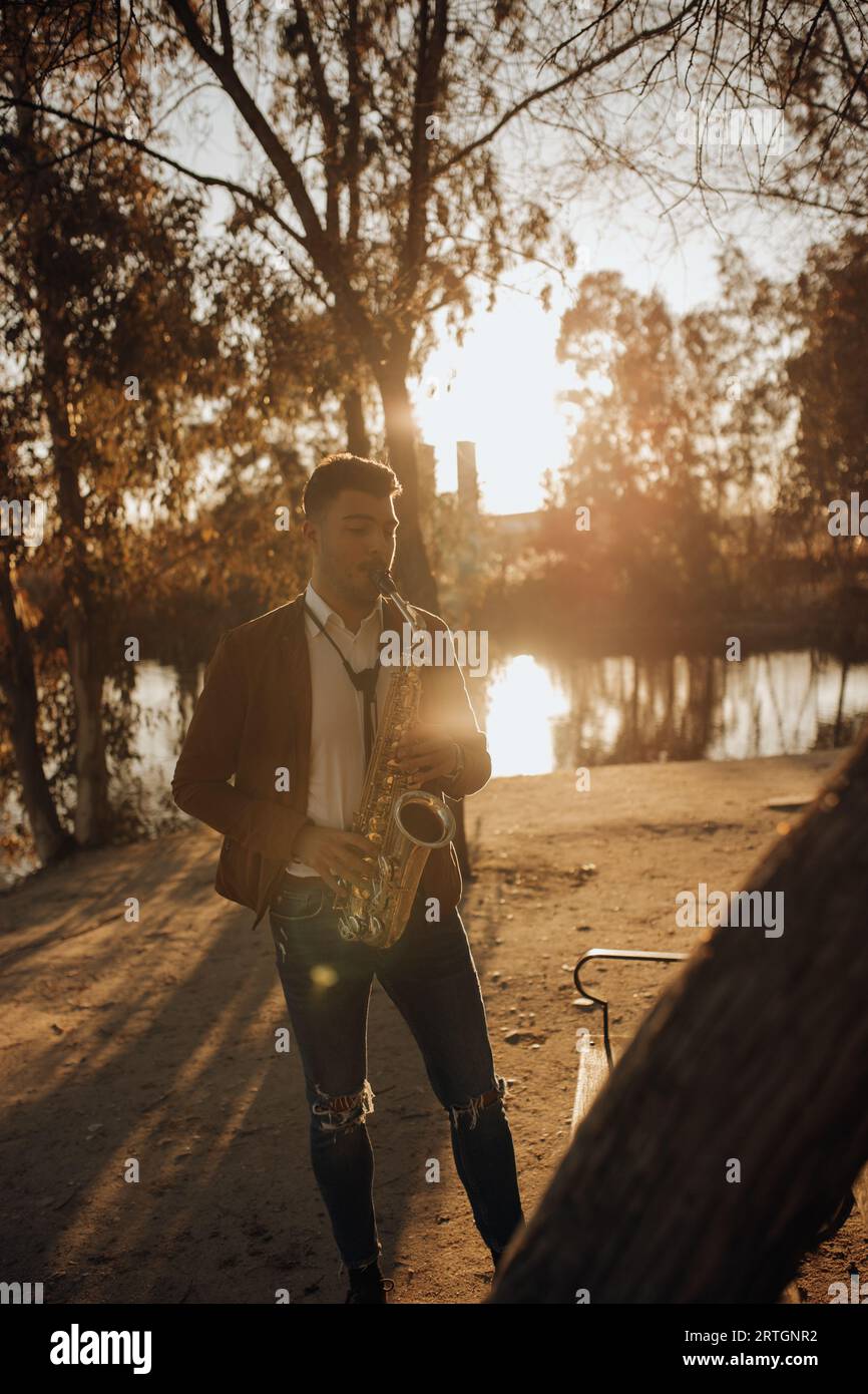 Young romantic musician playing on saxophone in park near water in ...