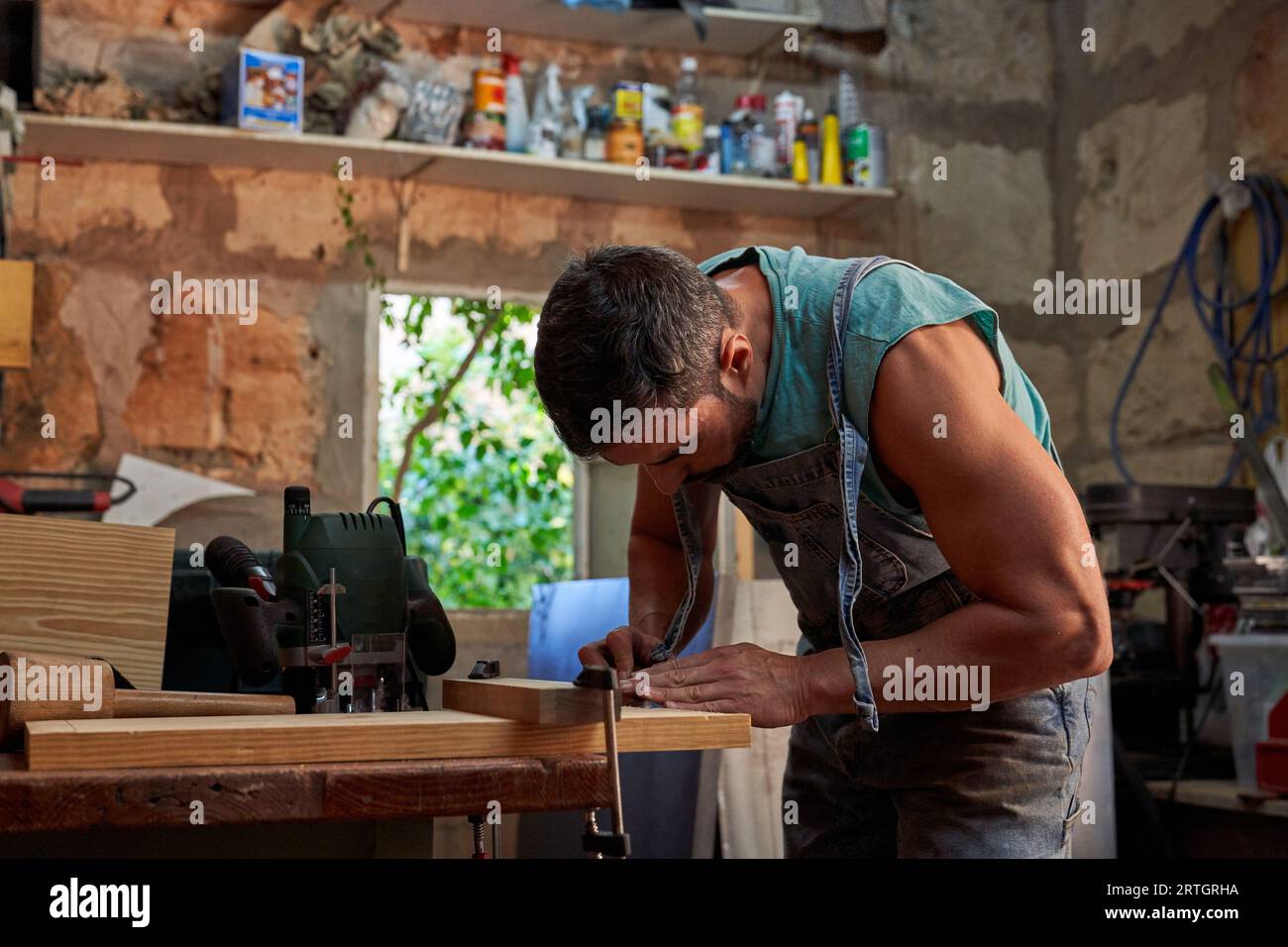 Side view of man wearing denim overall standing at workbench doing ...
