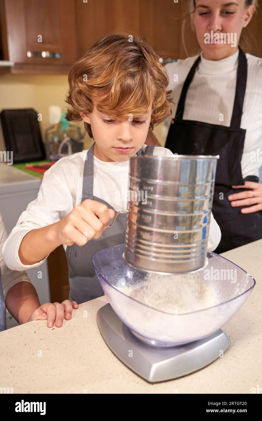High angle of focused boy in apron sifting flour in metal sieve to bowl ...