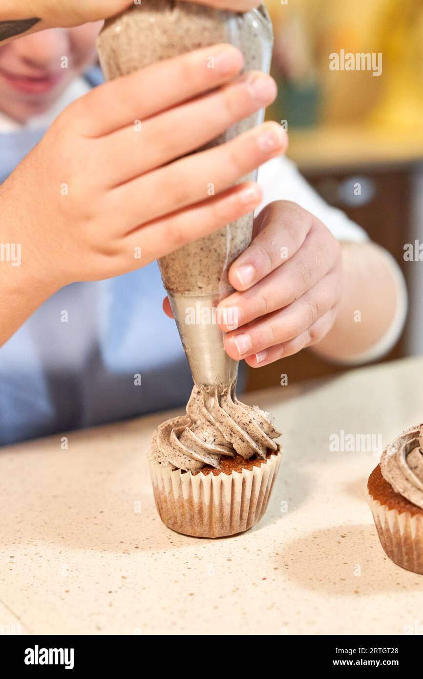 Crop anonymous kid in apron pouring cream on cupcake at table counter ...