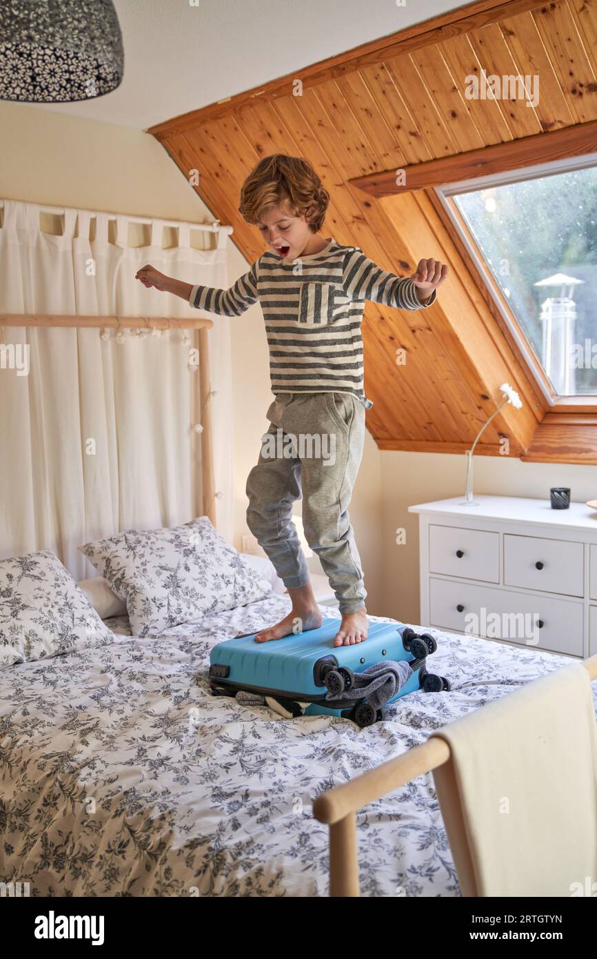 Full body of happy boy in casual clothes jumping on suitcase while ...