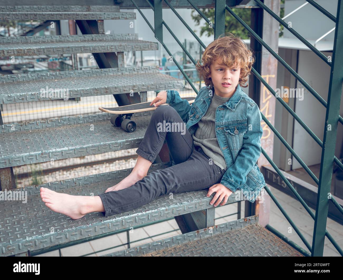 Full body of barefoot curly haired preteen boy in jeans jacket sitting on steps of staircase and ...