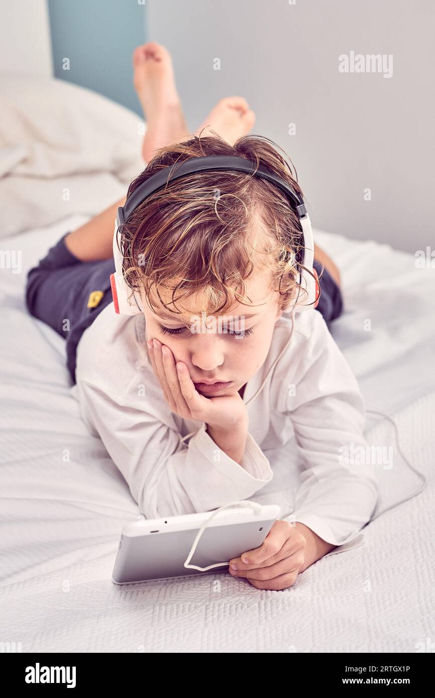 High angle of barefoot boy in headphones lying on bed and watching ...