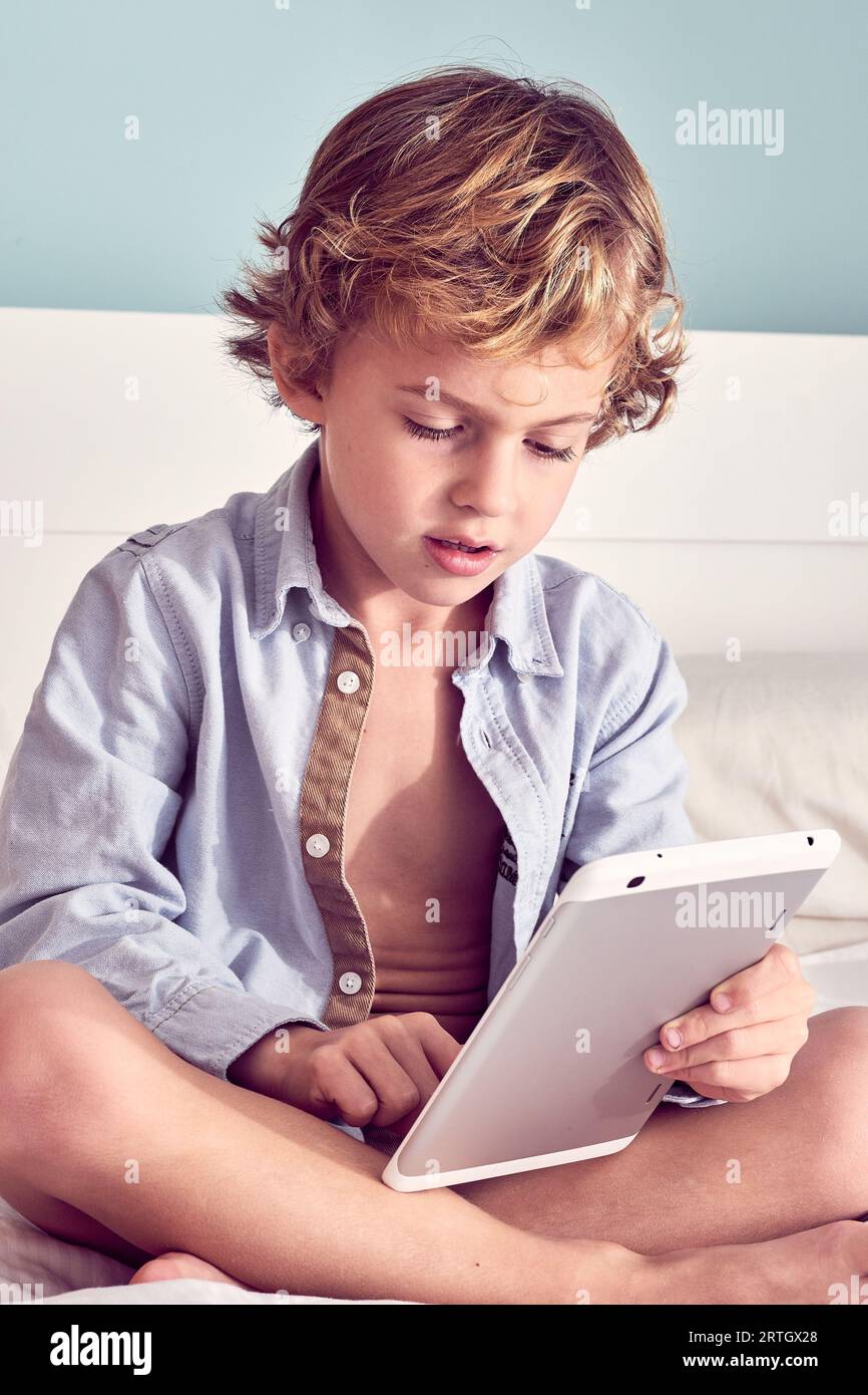 Cute boy wearing unbuttoned shirt sitting with crossed legs and playing ...