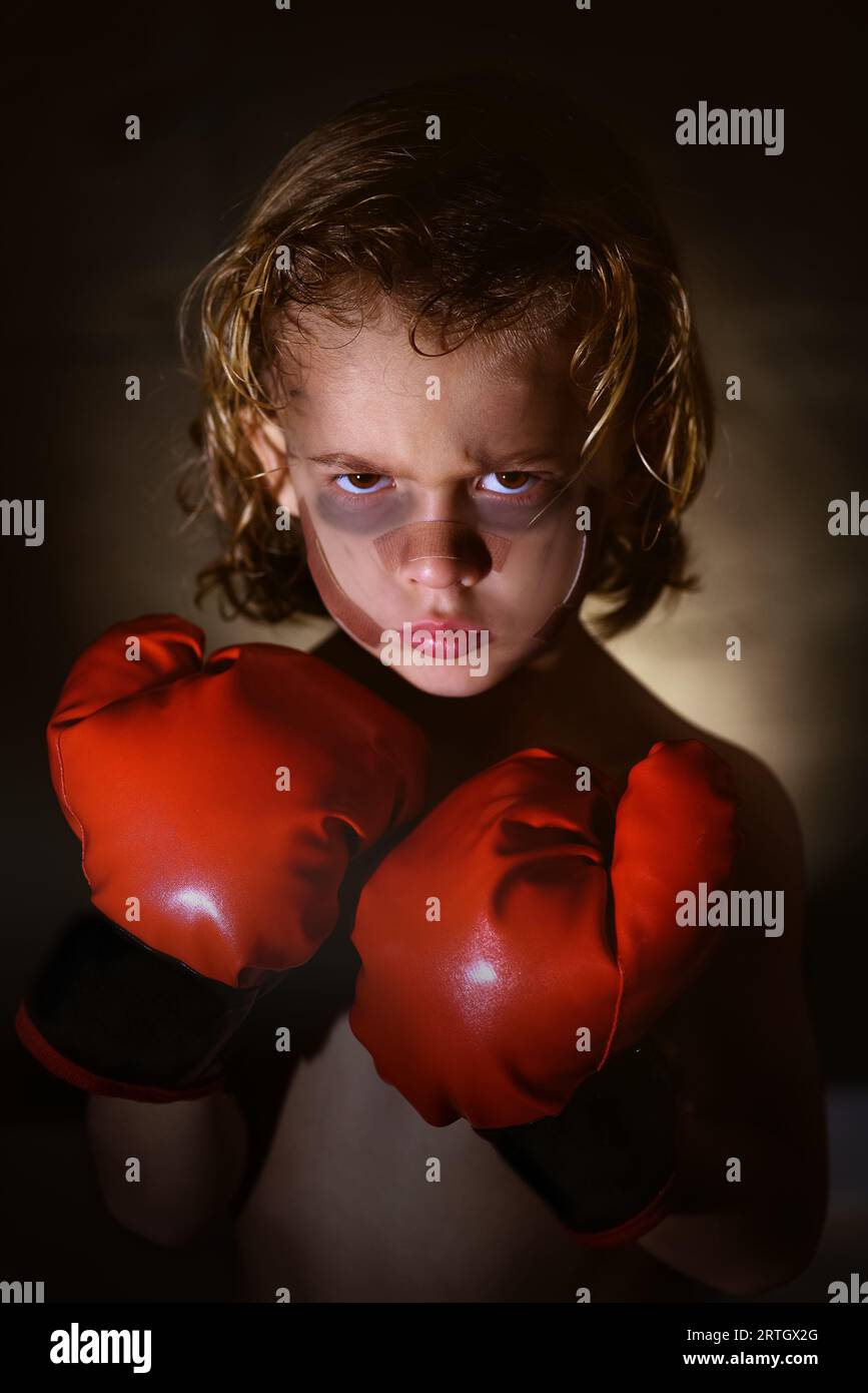 Serious boy with bruises and band aid on face in red boxing gloves ...