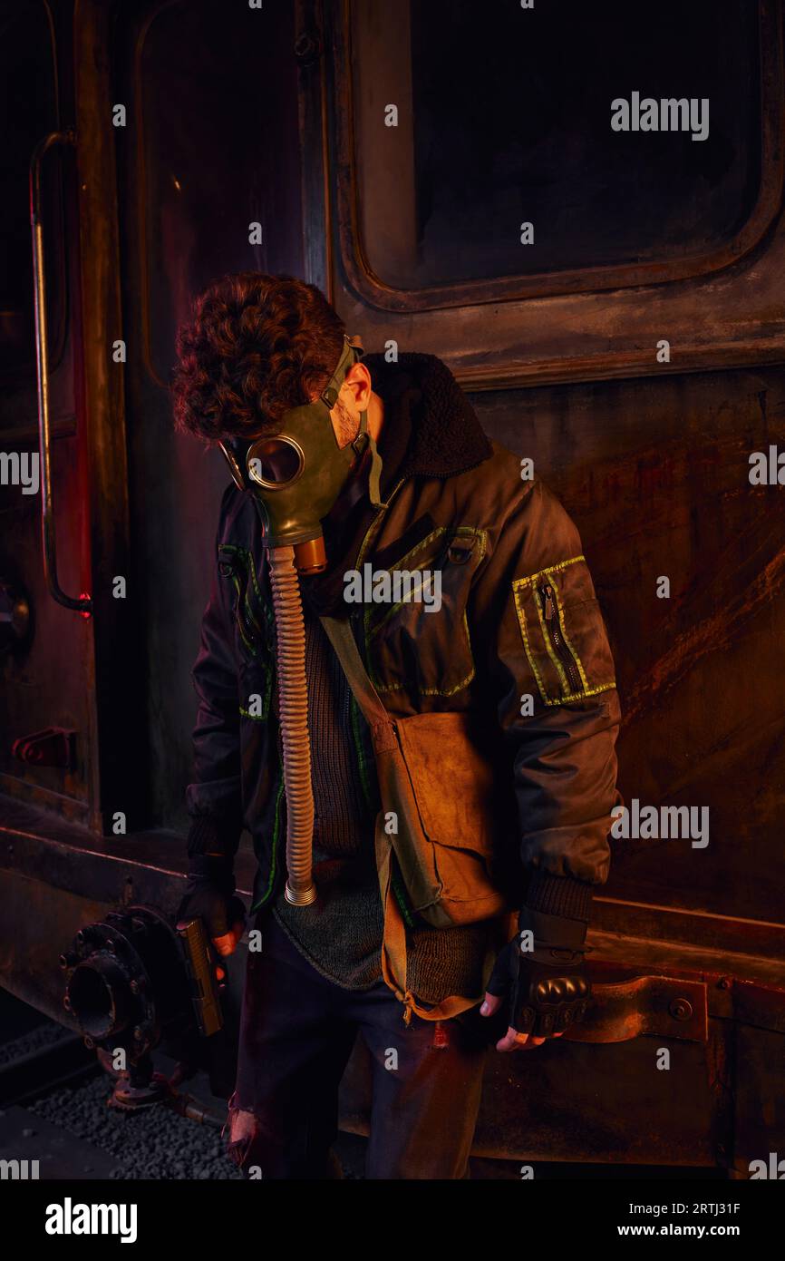 man in gas mask and worn outfit standing with gun near rusty carriage ...