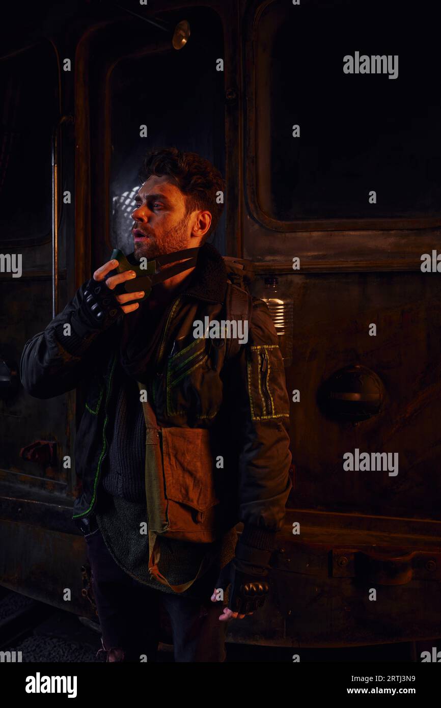 man in worn outfit holding gas mask and looking away in abandoned ...