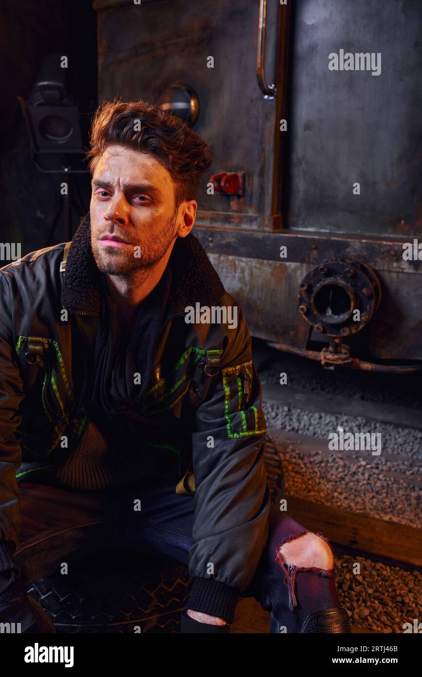 thoughtful unshaven man in worn jacket near rusty carriage in abandoned ...