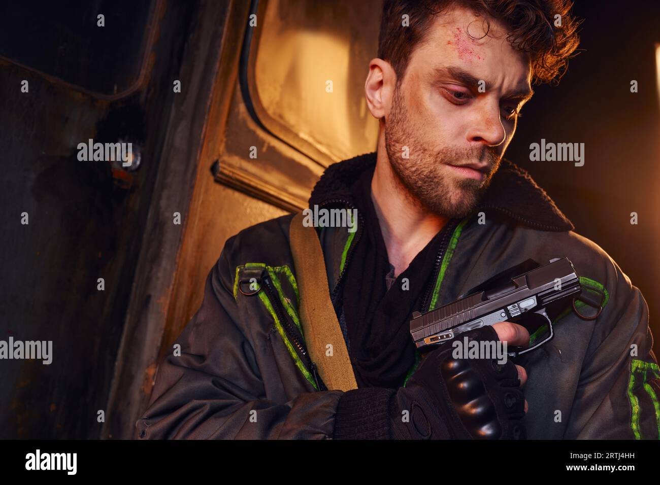 unshaven and gloomy man with scratched face looking at gun in dark ...