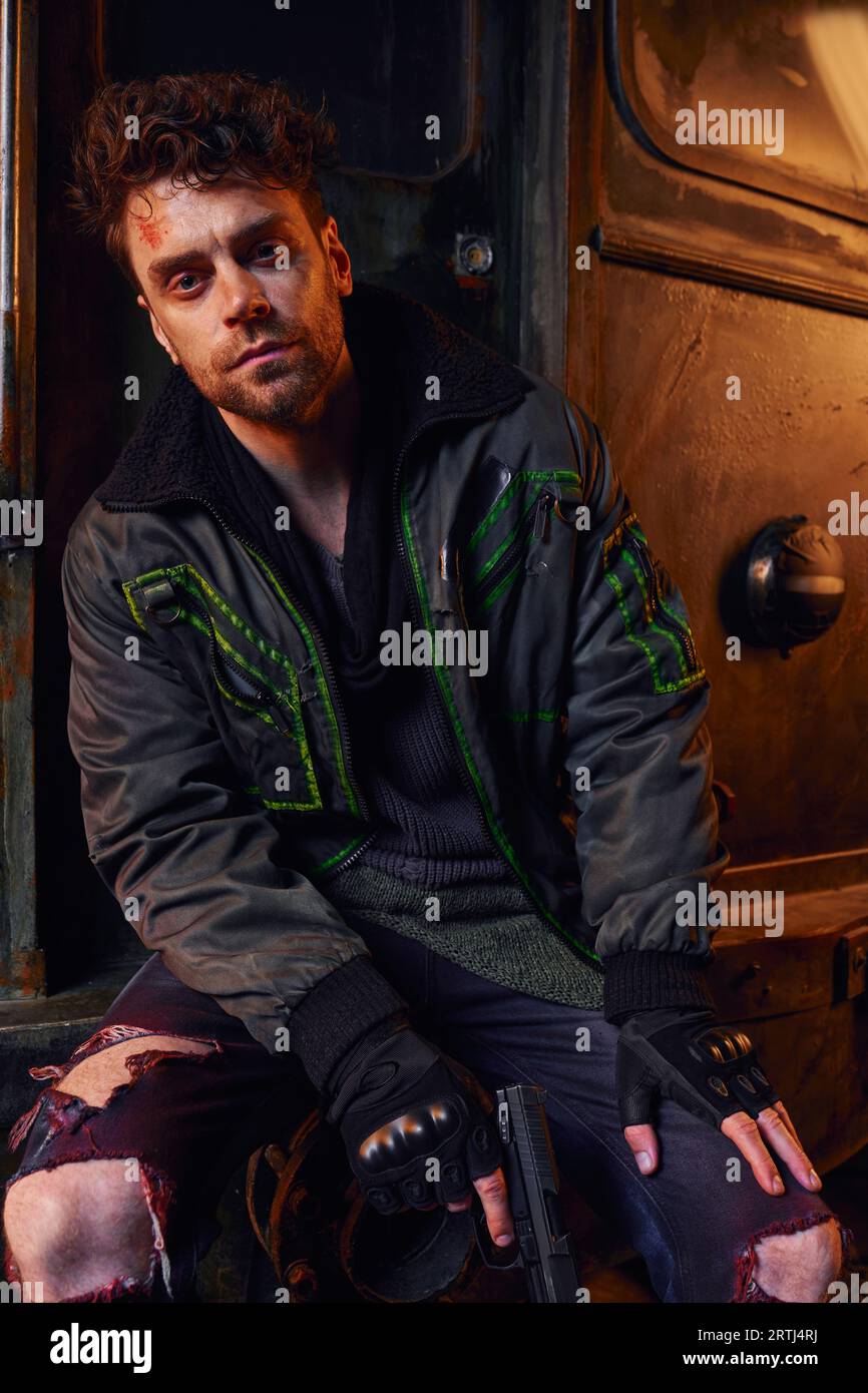 unshaven man in worn jacket holding gun and looking at camera in ...