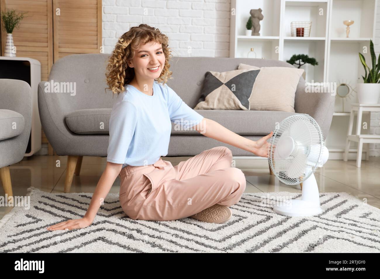 Beautiful happy young woman with electric fan sitting on floor in ...