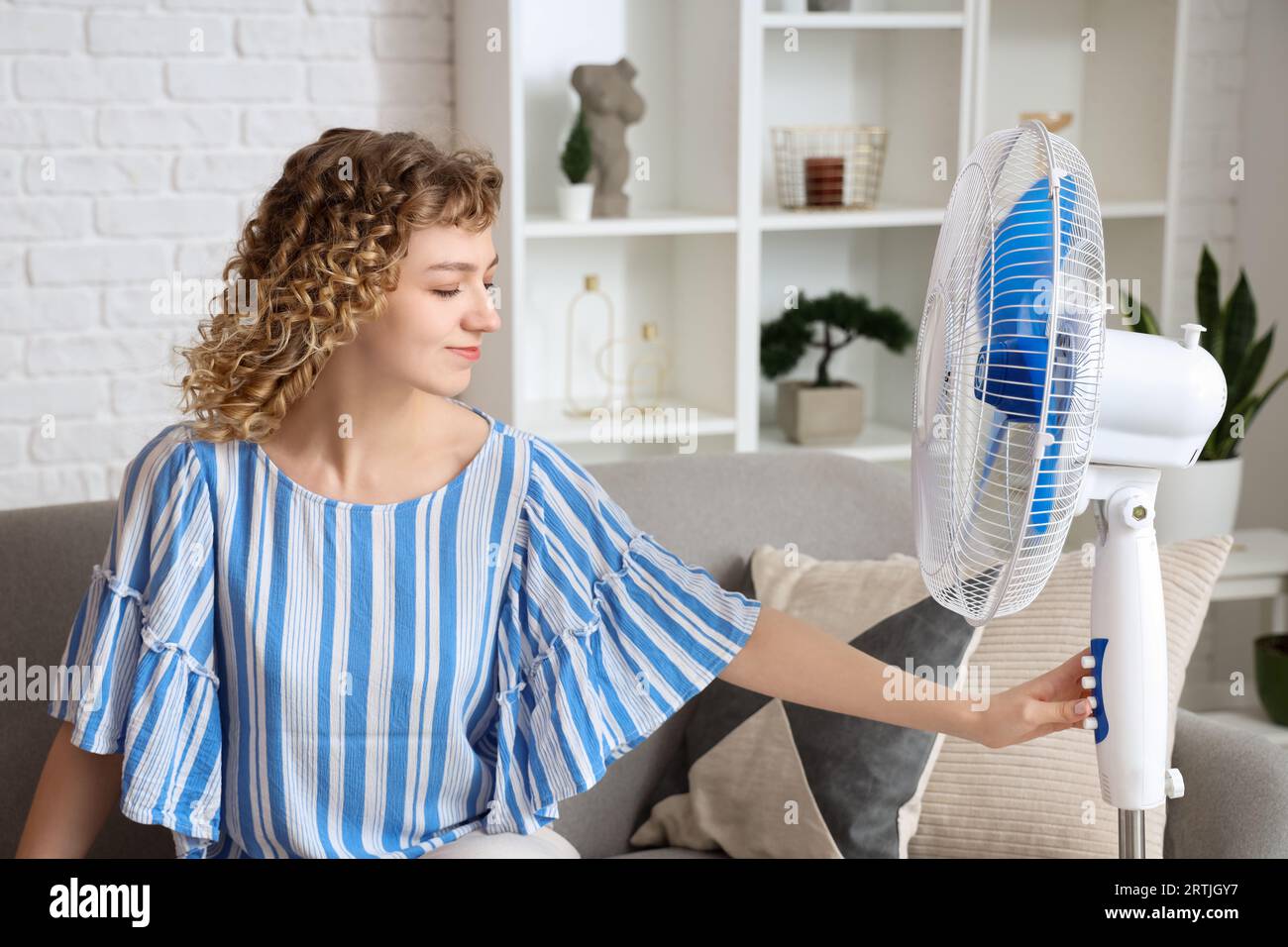 Beautiful happy young woman with electric fan sitting on sofa in living ...