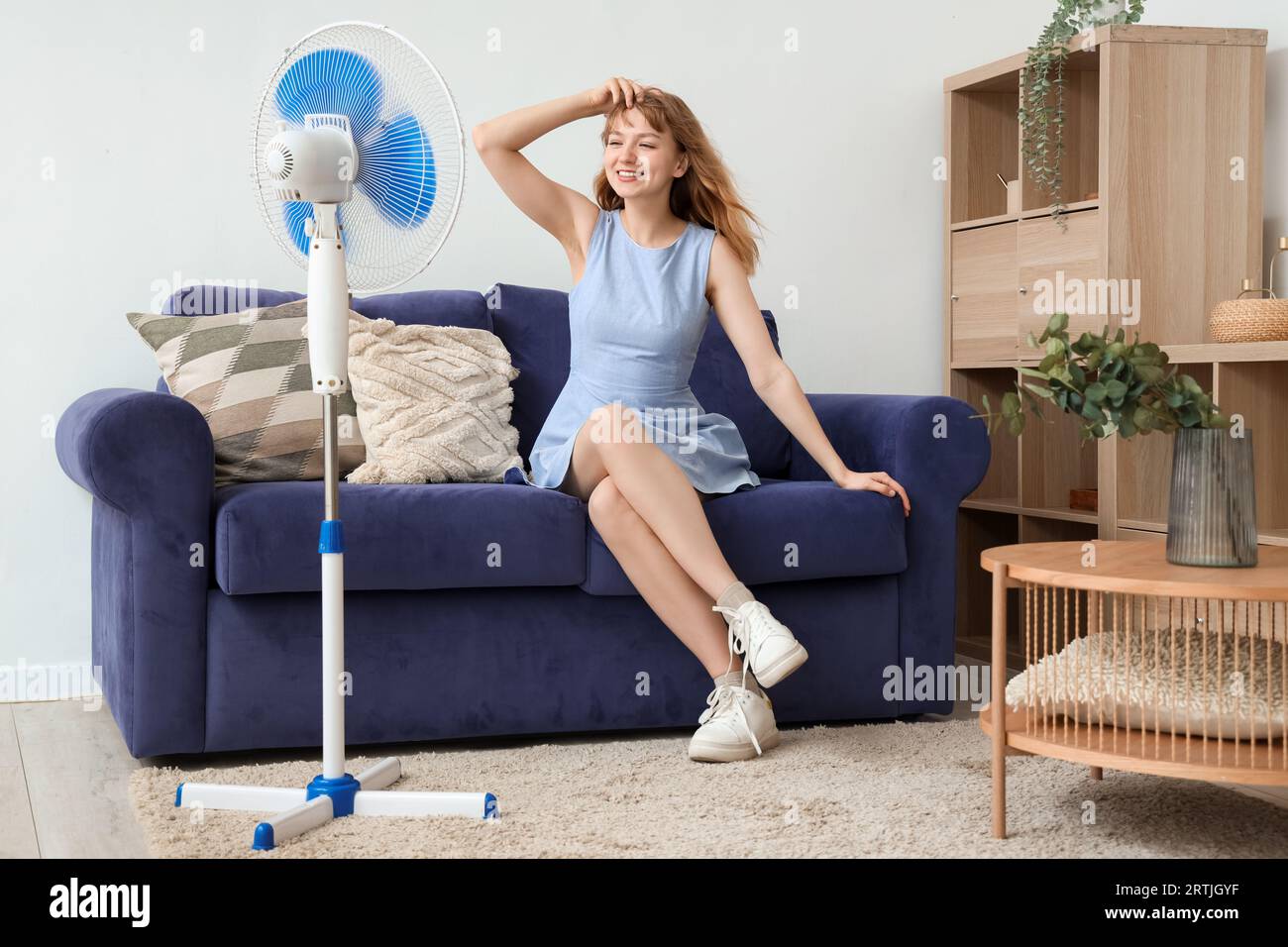 Happy beautiful young woman with electric fan sitting on sofa in living ...
