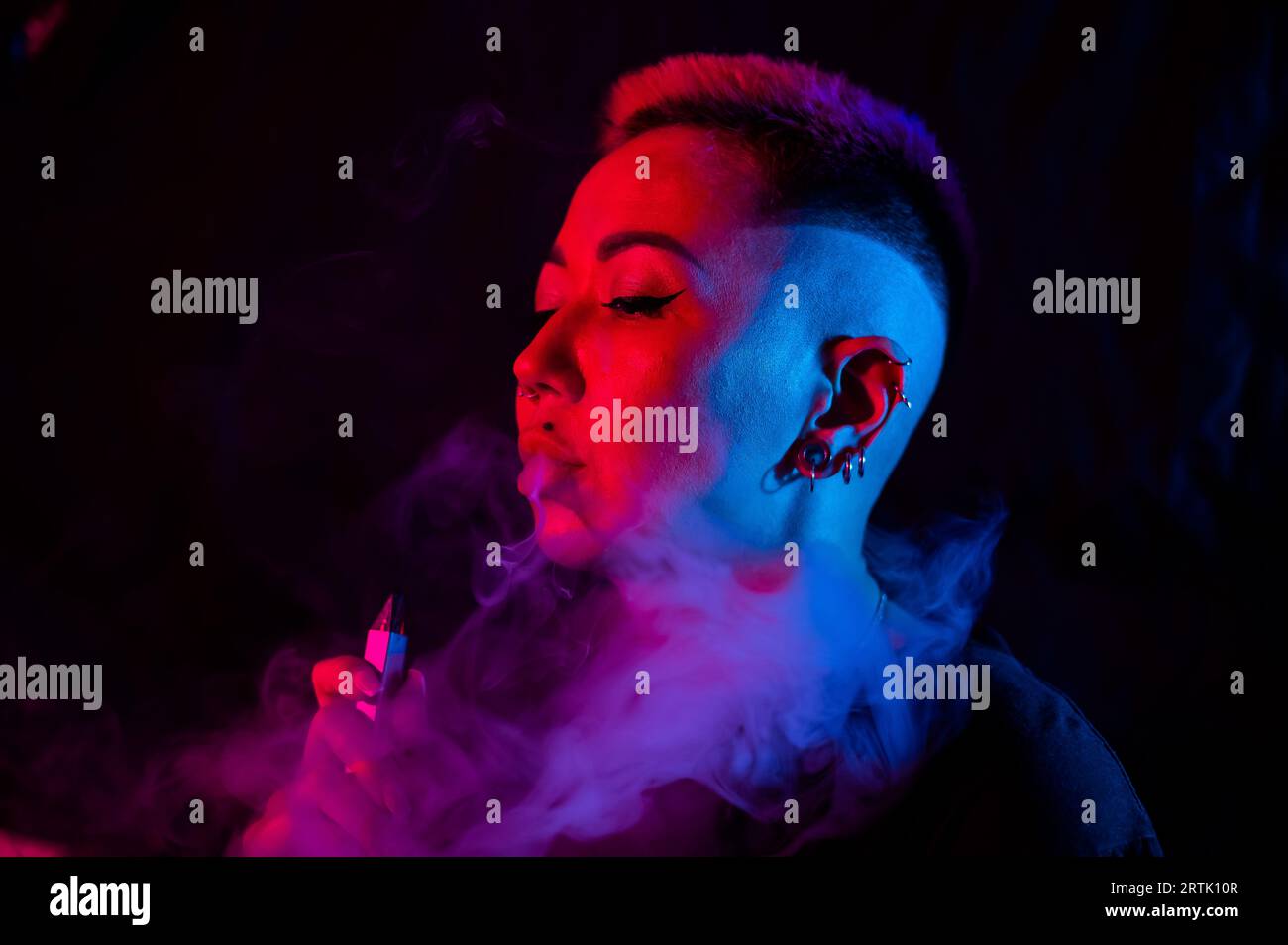 Portrait asian woman with shaved temples smokes vape in neon light ...