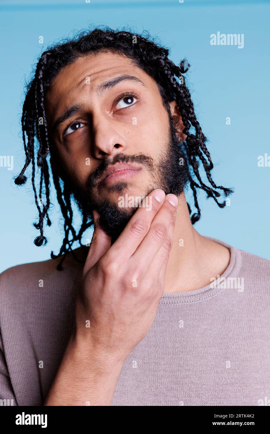 Arab young adult man rubbing chin in contemplation with puzzled facial ...