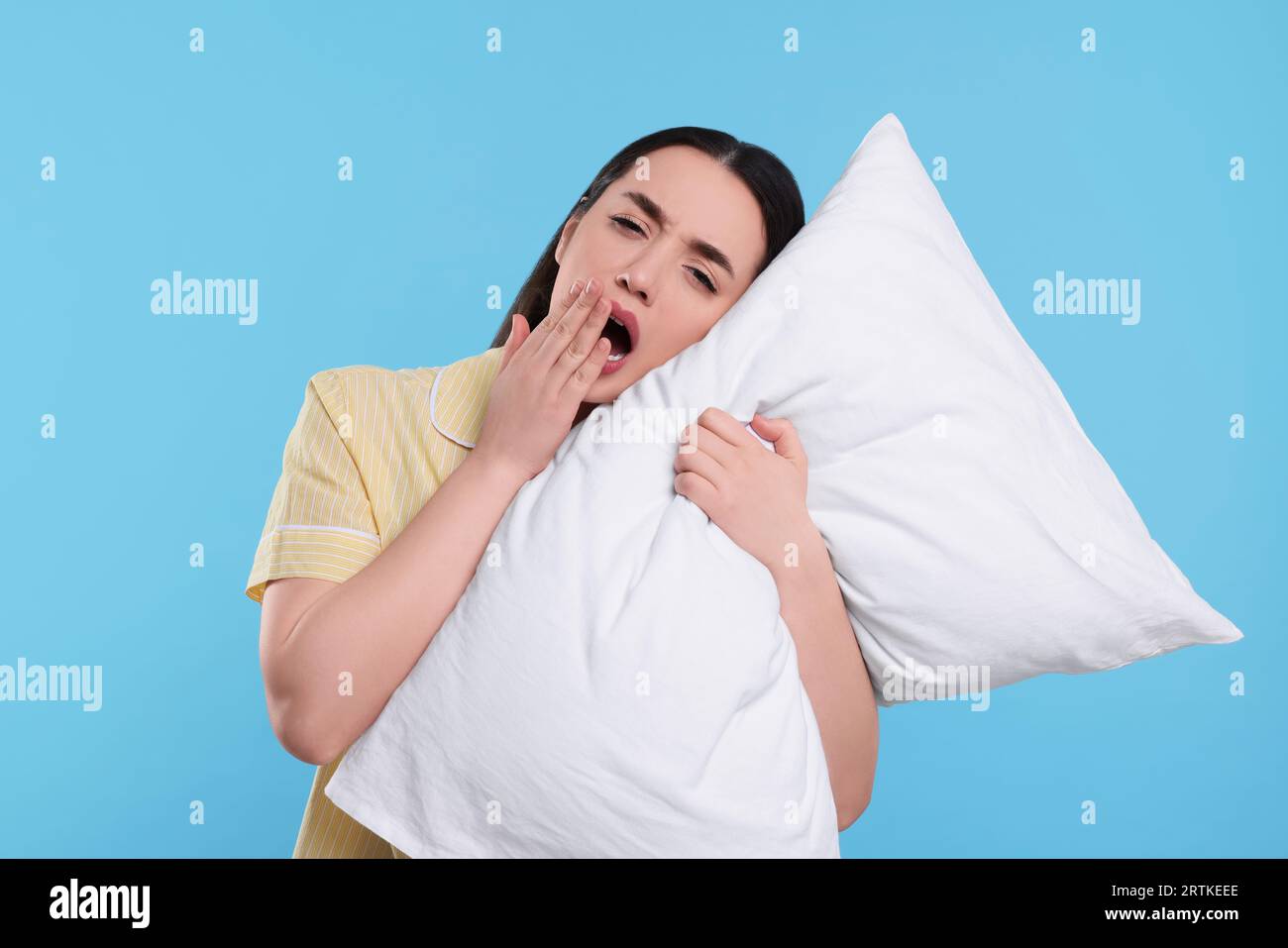 Tired young woman with pillow yawning on light blue background ...
